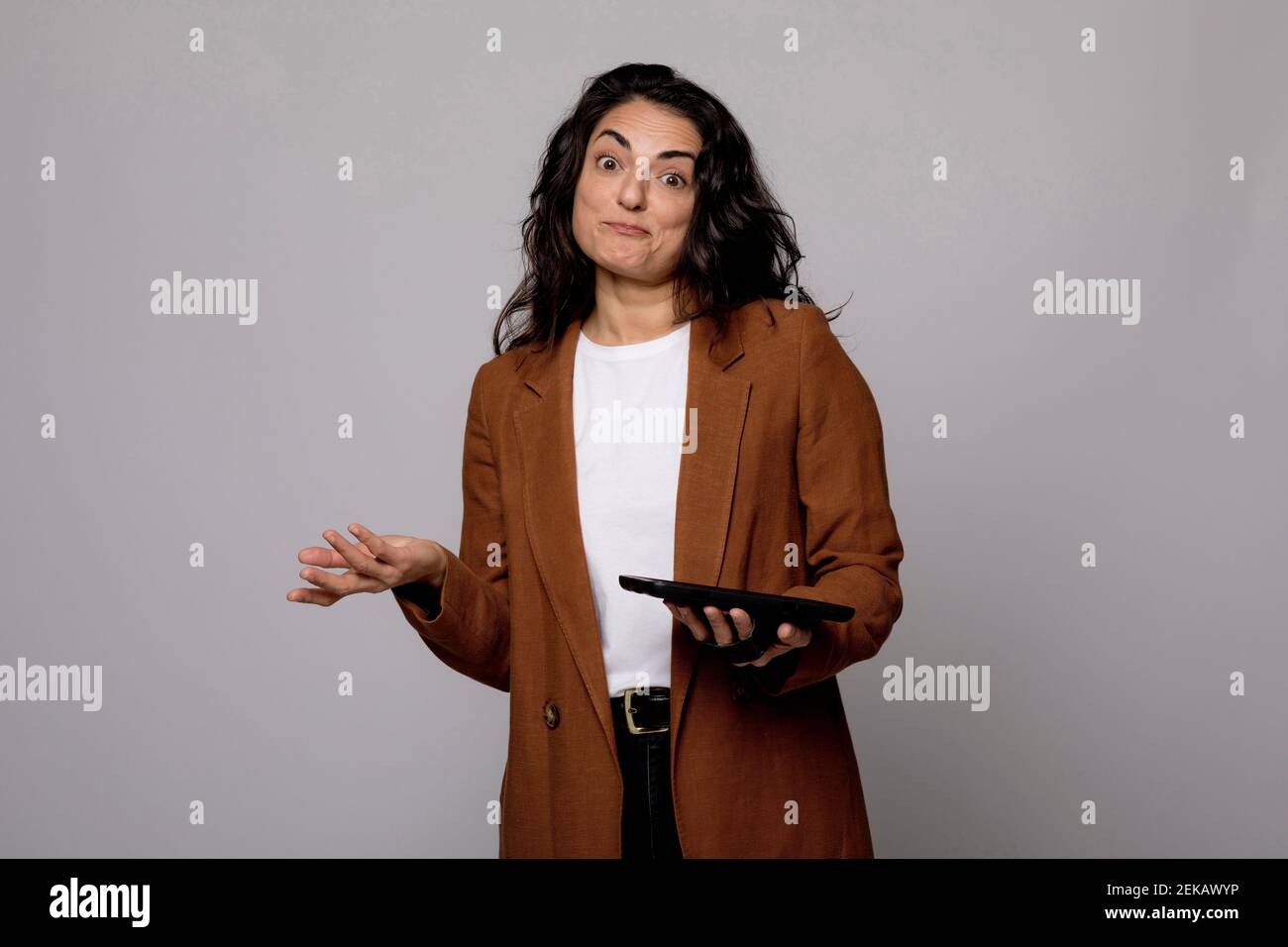 Confused woman with digital tablet gesturing against gray background ...