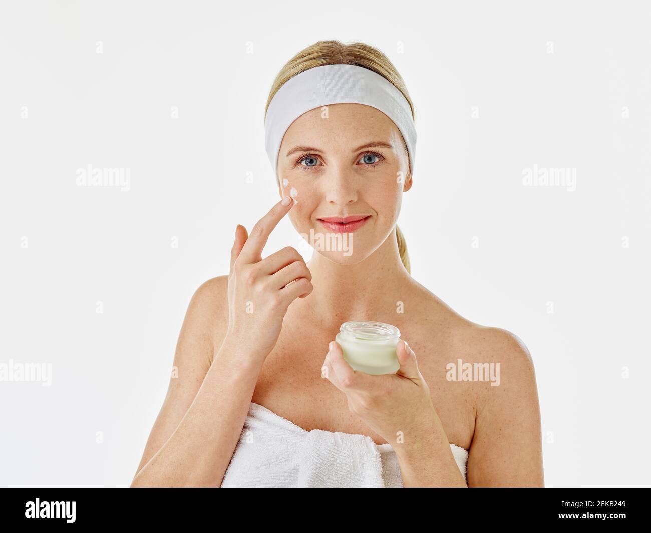 Woman wearing headband and towel applying face cream while standing ...