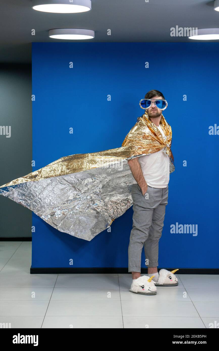 Businessman in gold color wrapping paper cape looking away by blue wall ...