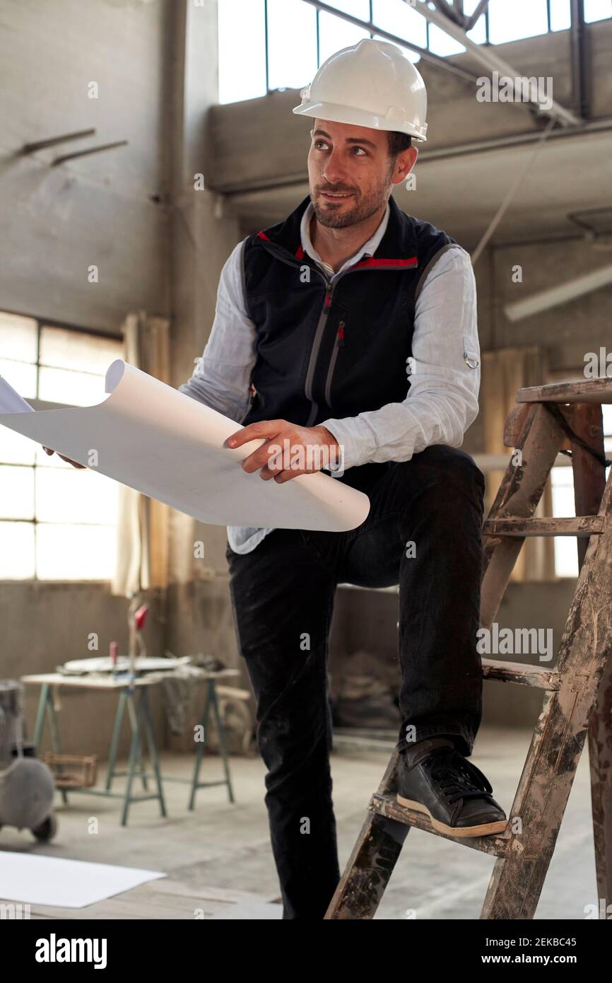 Male engineer holding blueprint looking away while standing on ladder ...