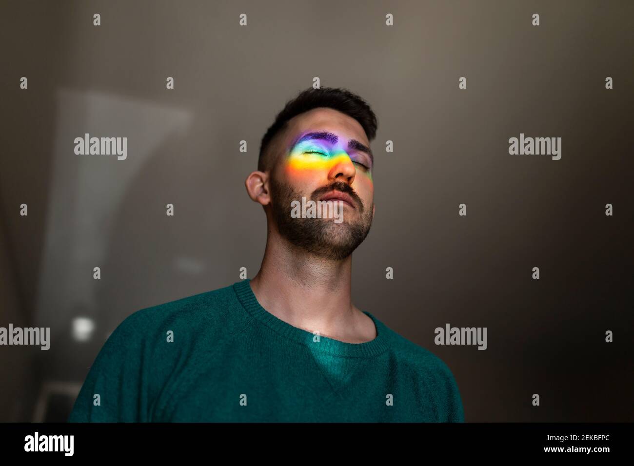 Rainbow light hitting face of young man standing indoors with closed ...