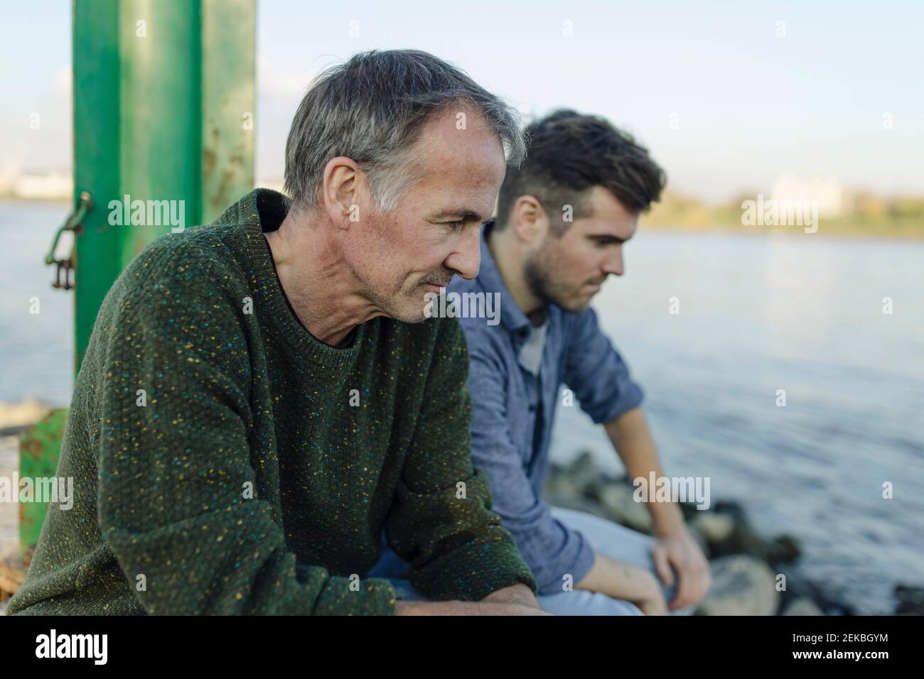 Son and father looking down while sitting by river Stock Photo - Alamy