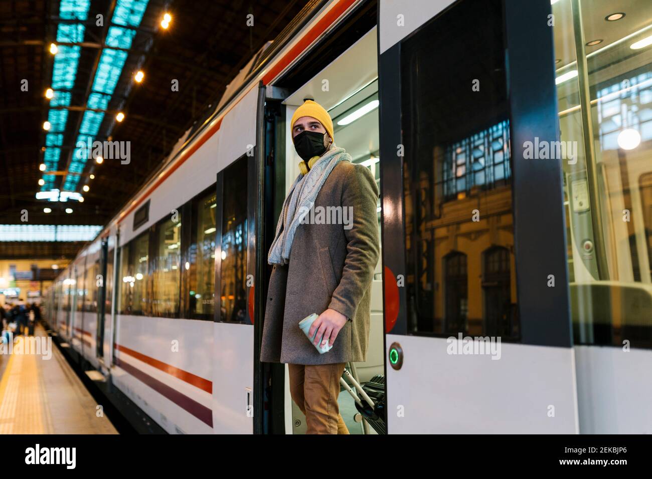 Mid adult man wearing face mask stepping out of train with coffee cup ...