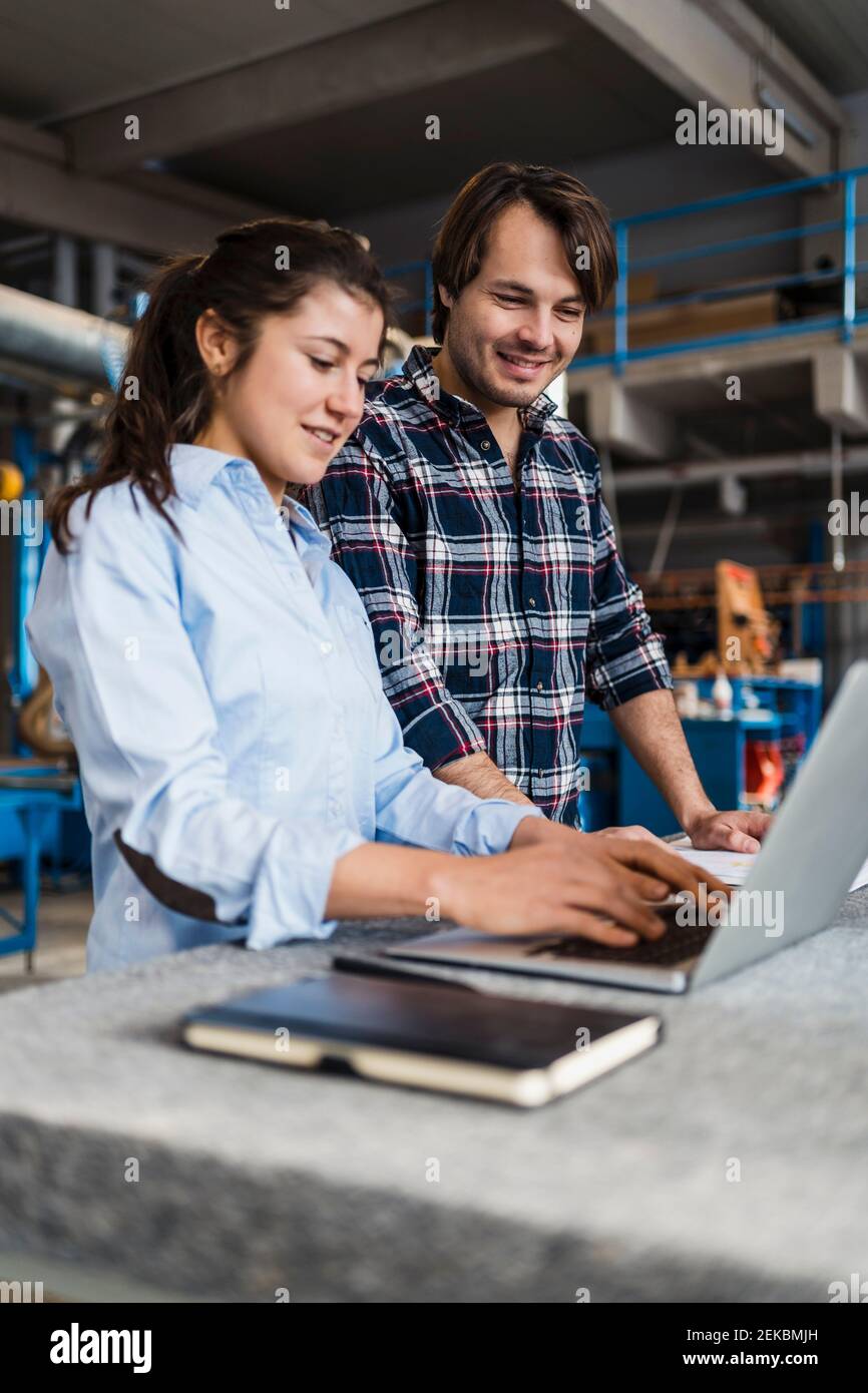 Smiling business people working on laptop while standing at industry ...