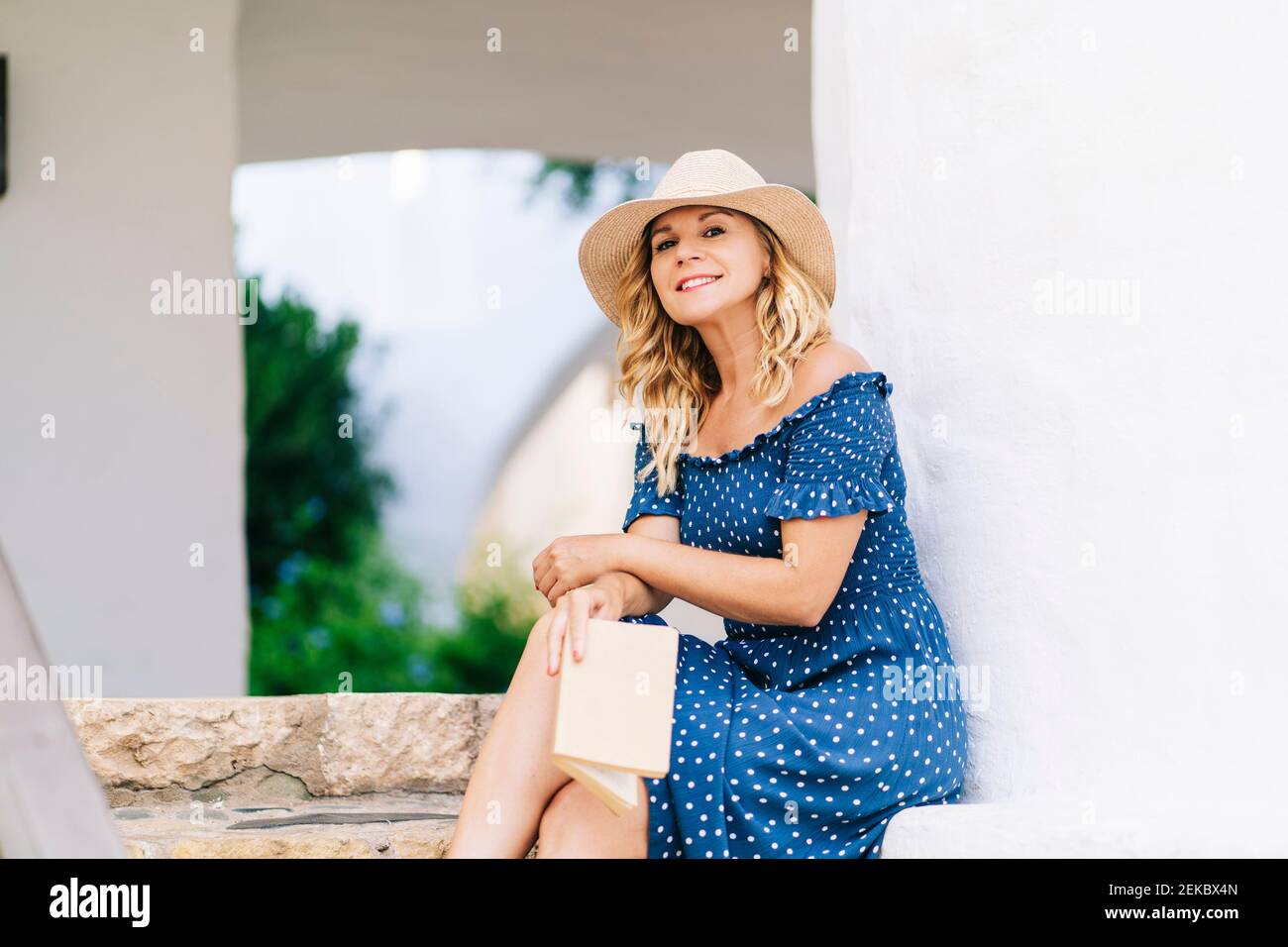 Smiling beautiful woman wearing hat holding book while sitting against ...