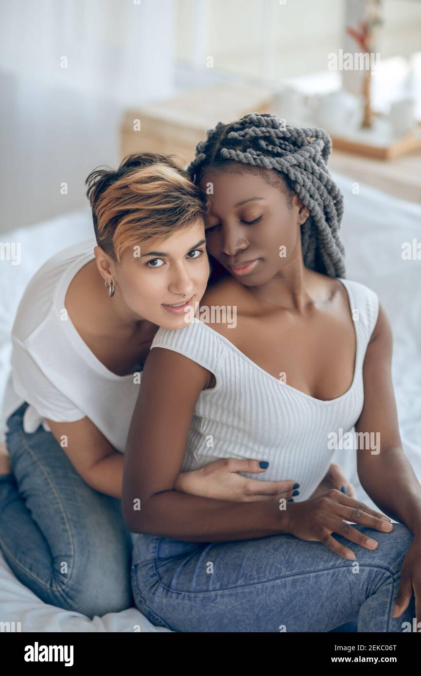 Short Haired Girl Hugging Her Dark Skinned Girlfriend From The Back And Looking Pleased Stock