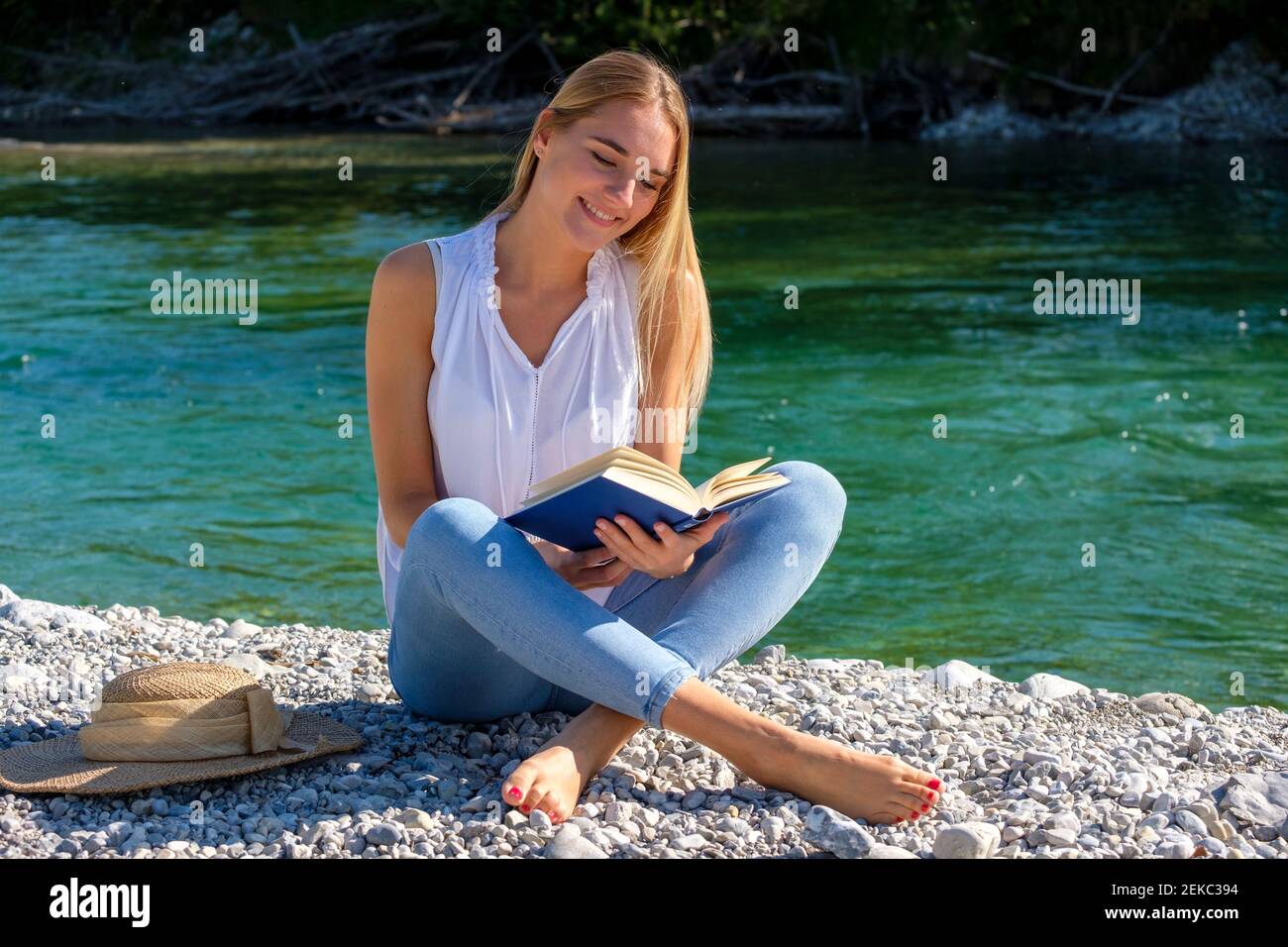 Smiling blond woman sitting cross-legged while reading book by river ...