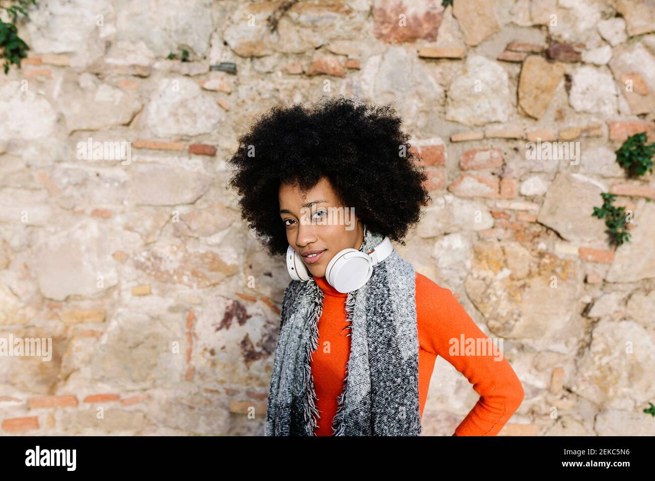 Young woman with headphones and scarf standing against stone wall on ...