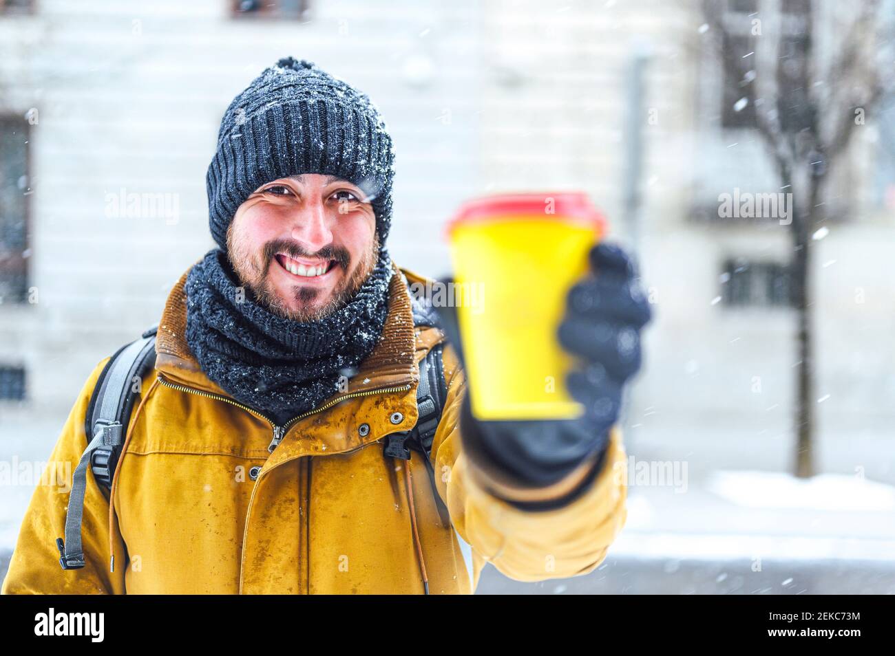 Happy man showing coffee cup at camera while snowing during winter ...