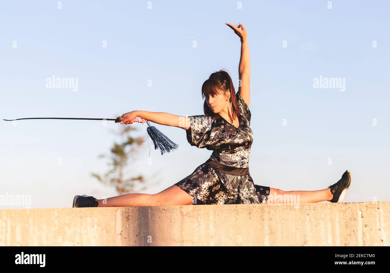 Young female athlete holding sword while doing split on built structure ...