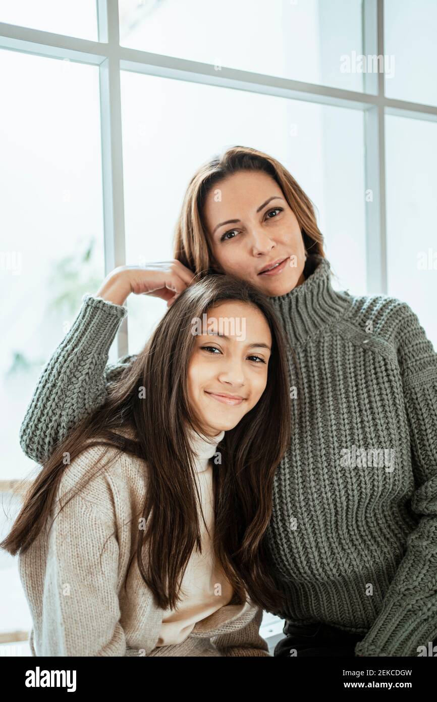 Smiling mother and daughter sitting against glass window at home Stock Photo - Alamy