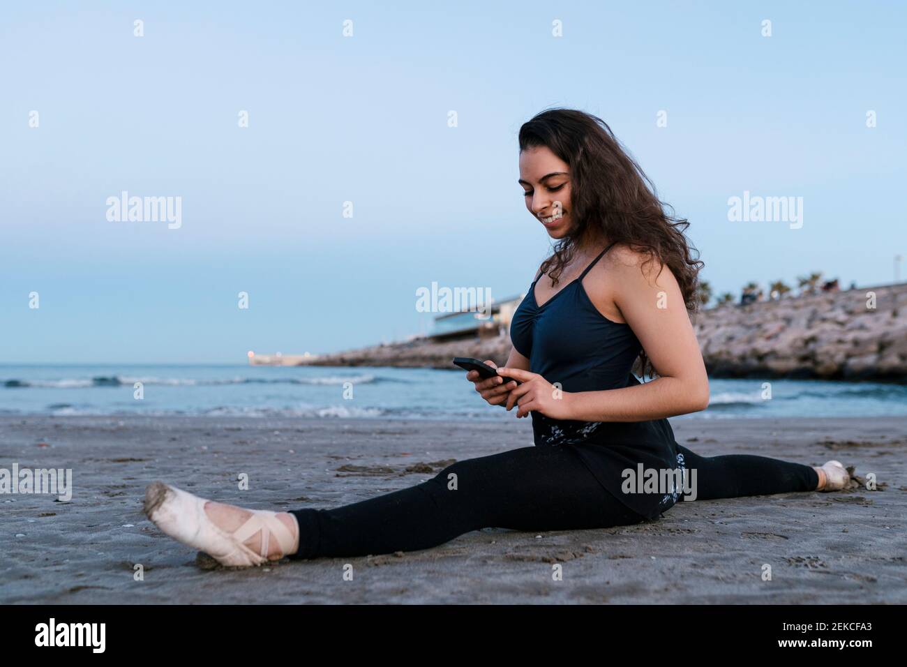 Flexible female dancer using mobile phone while doing splits at beach ...