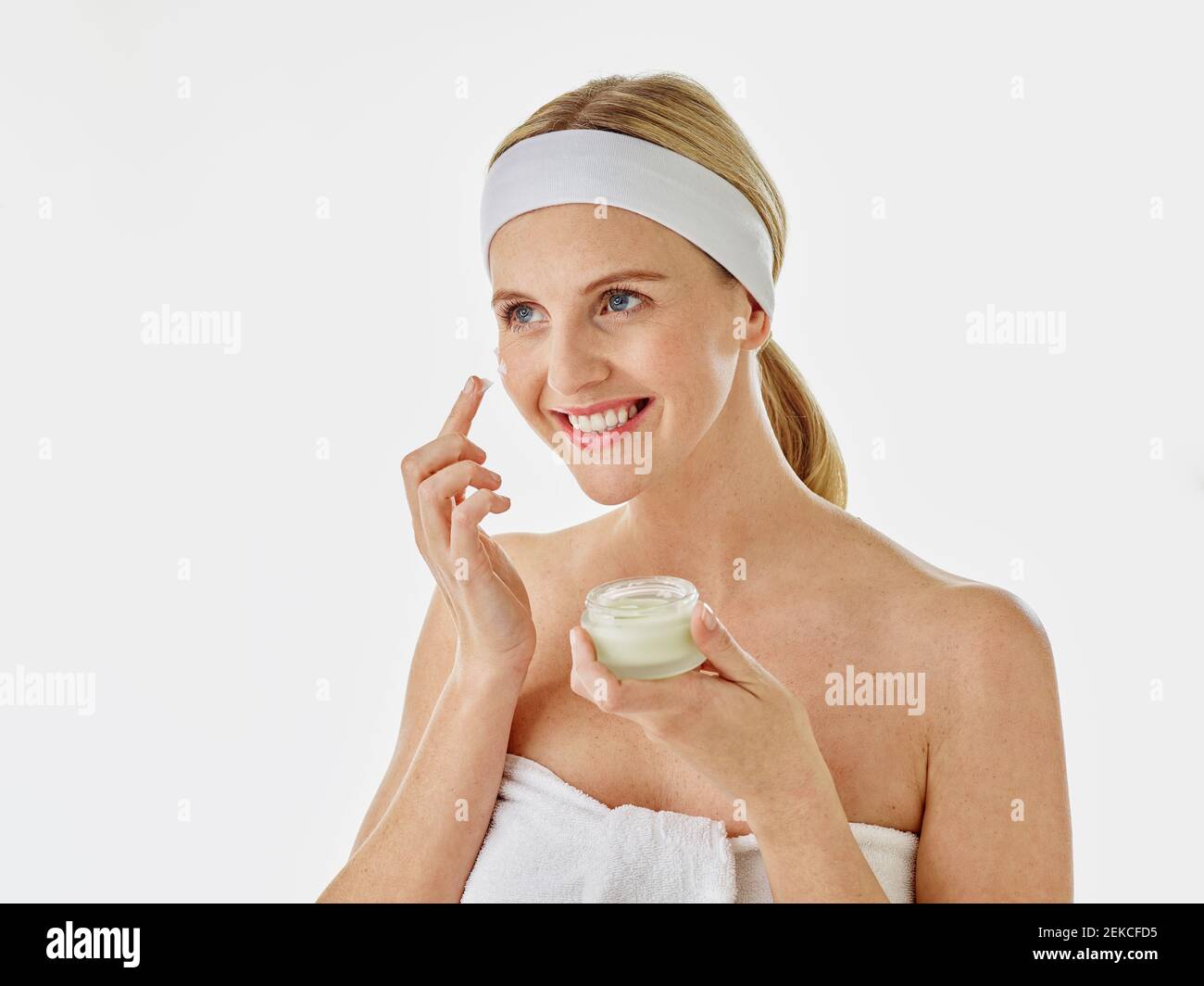 Young woman smiling while applying facial cream standing against white ...
