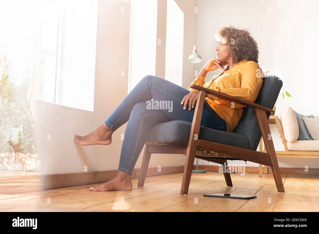 Thoughtful woman with hand on chin looking through window while sitting ...