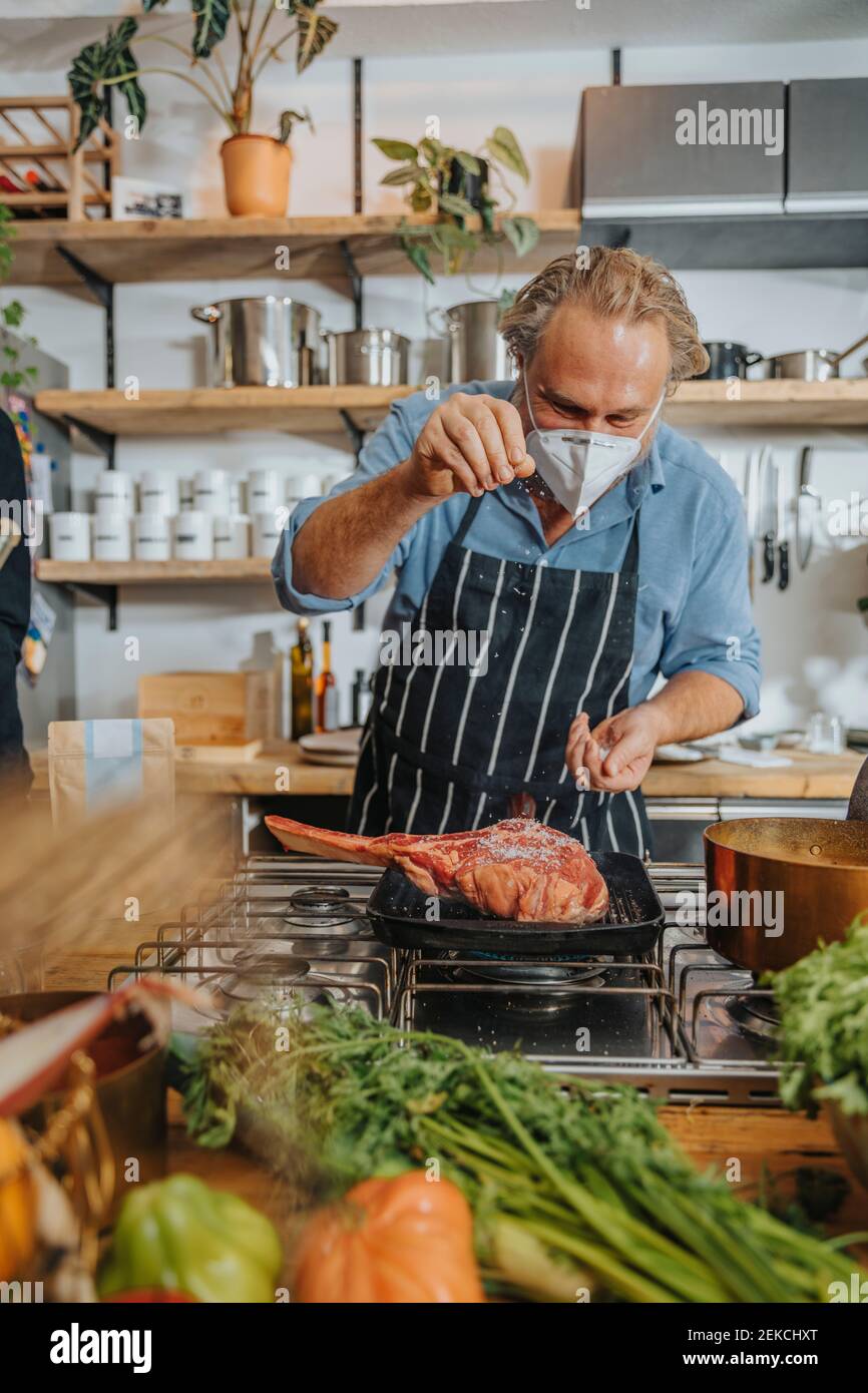 Mature chef wearing protective face mask adding seasoning on tomahawk ...