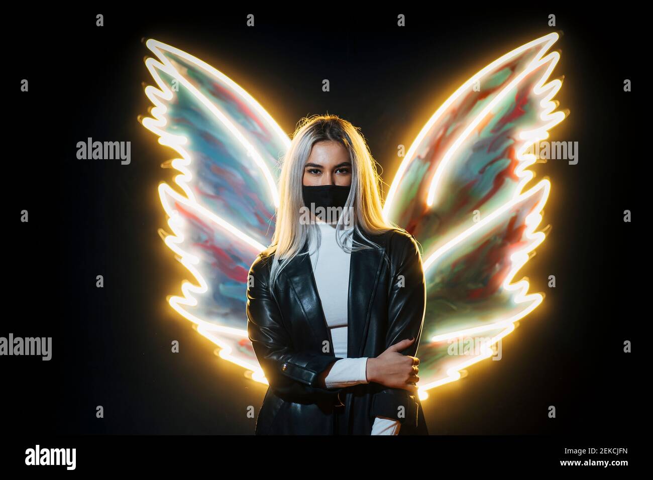Young woman wearing face mask posing with illuminated wings against ...