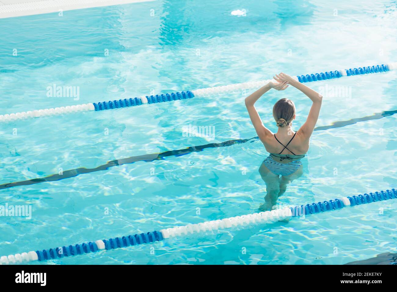 Back view of young woman standing with raised hands in swimming pool ...