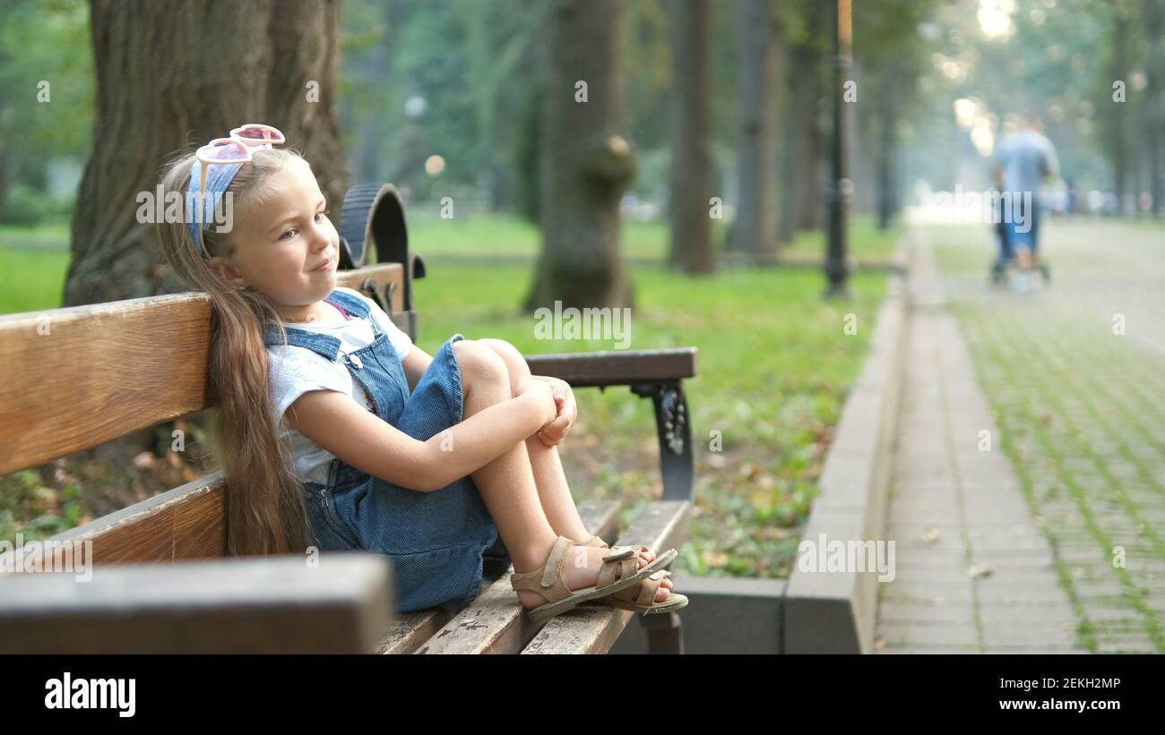 Small irritated child girl sitting alone on a bench in summer park ...