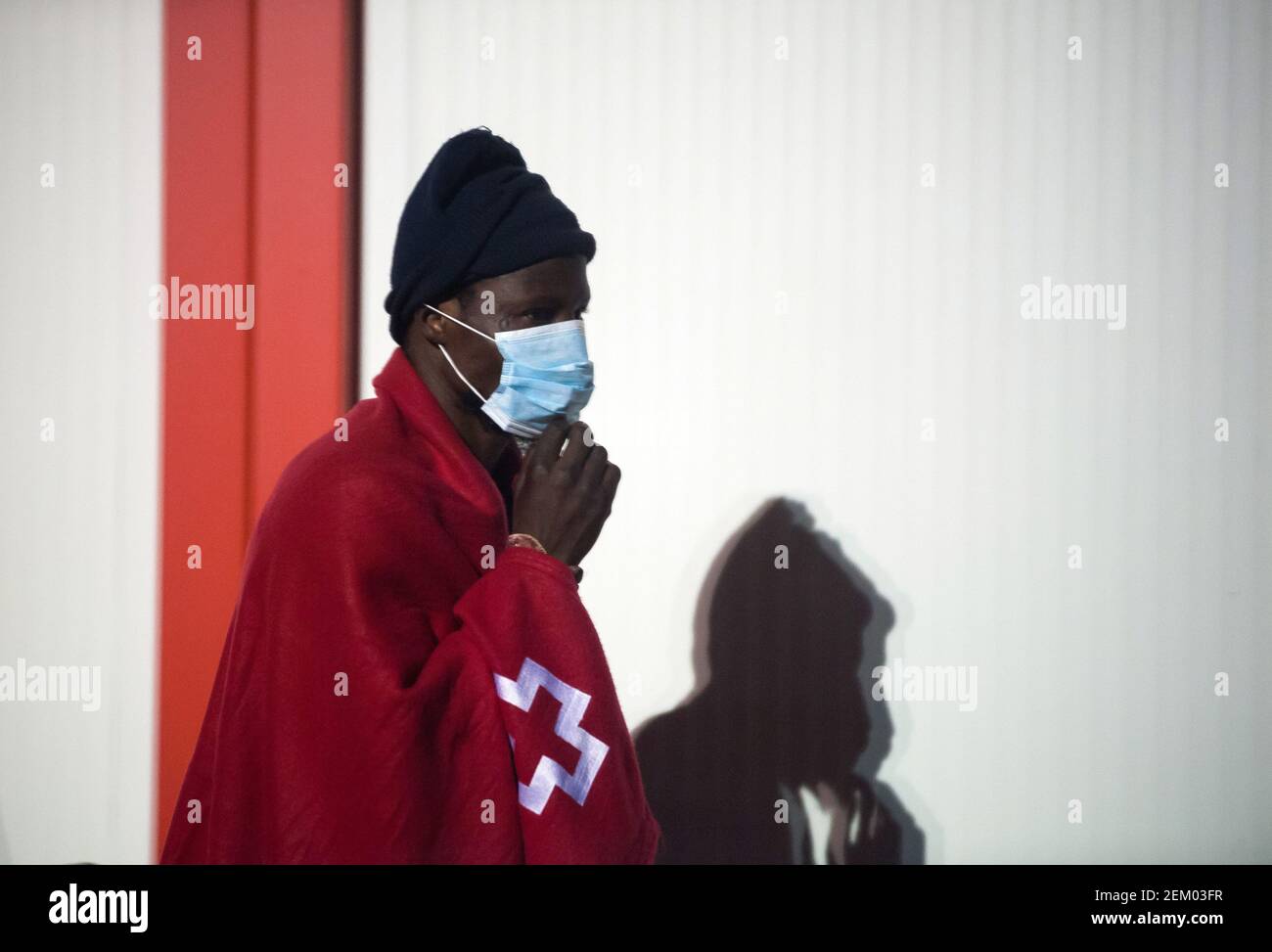 A migrant wearing a face mask is wrapped with a red blanket as he walks ...