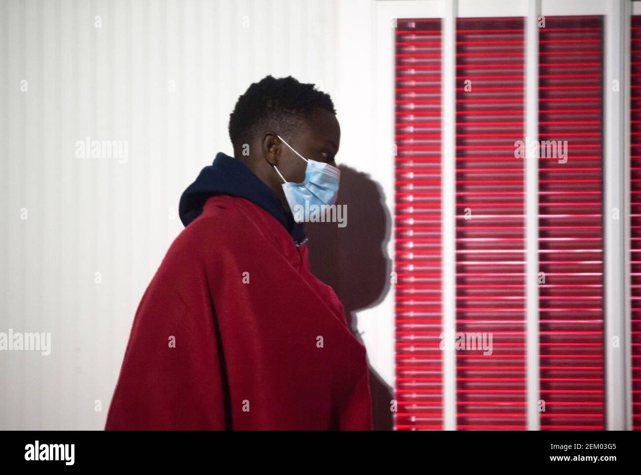 A migrant wearing a face mask is wrapped with a red blanket as he walks ...