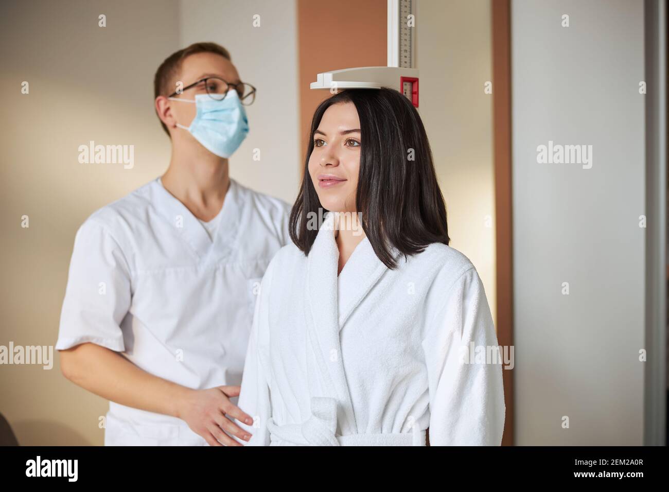 Serious bespectacled physician in a face mask applying a stadiometer ...