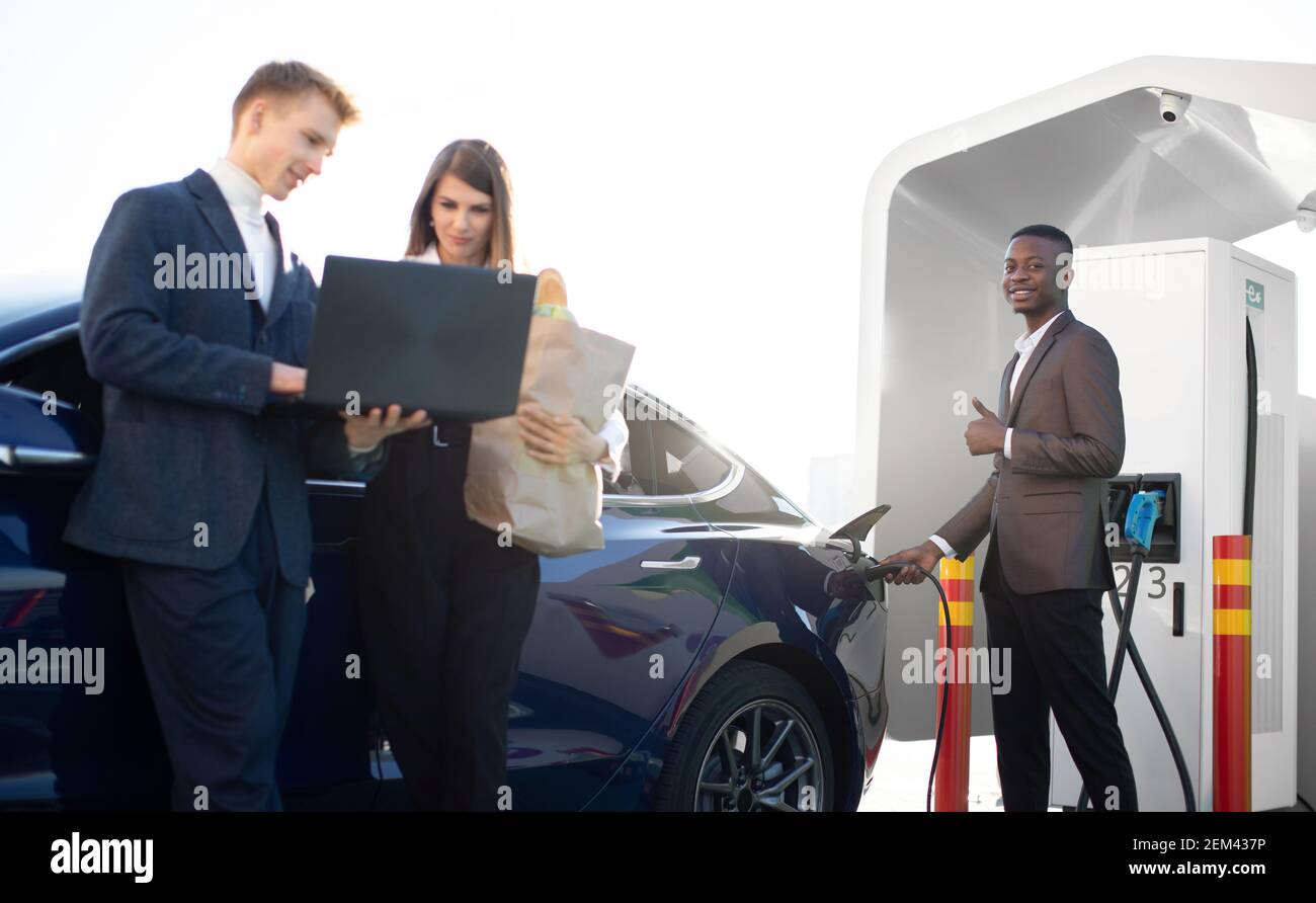 Smiling African man in formal wear, inserting plug into electric car ...
