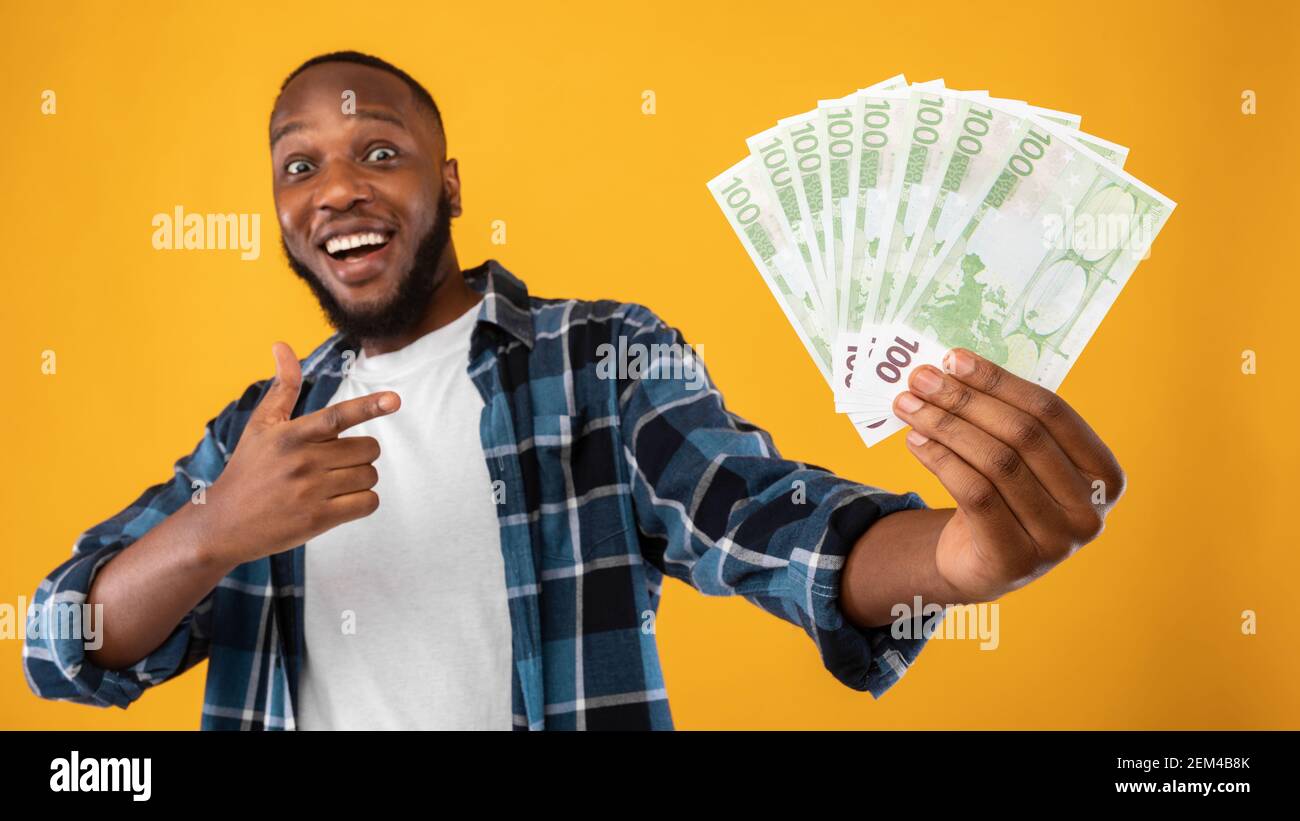 African Man Holding Money Pointing Finger At Cash, Yellow Background ...