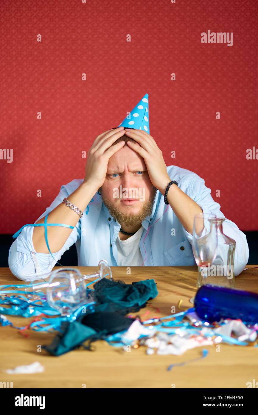 Man suffering from hangover at table with blue cap in messy room after ...