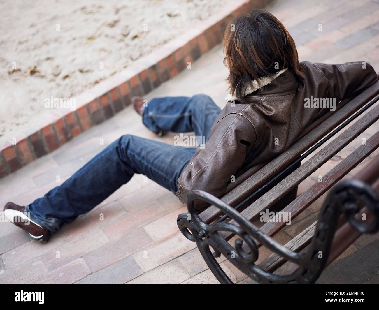 Rear view of a person sitting on ground and leaning against a bench ...