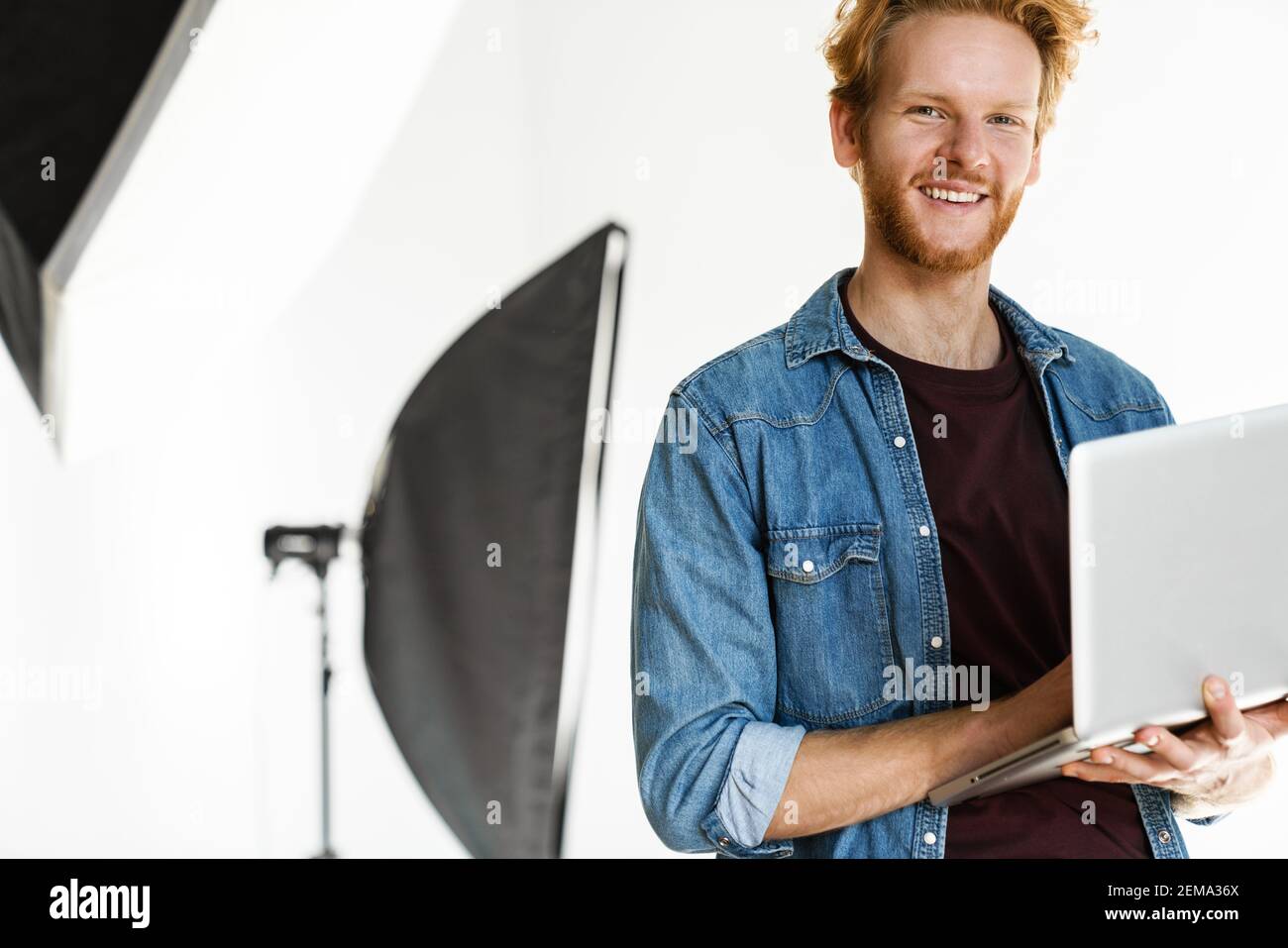 Happy handsome ginger guy smiling while working with laptop in studio ...