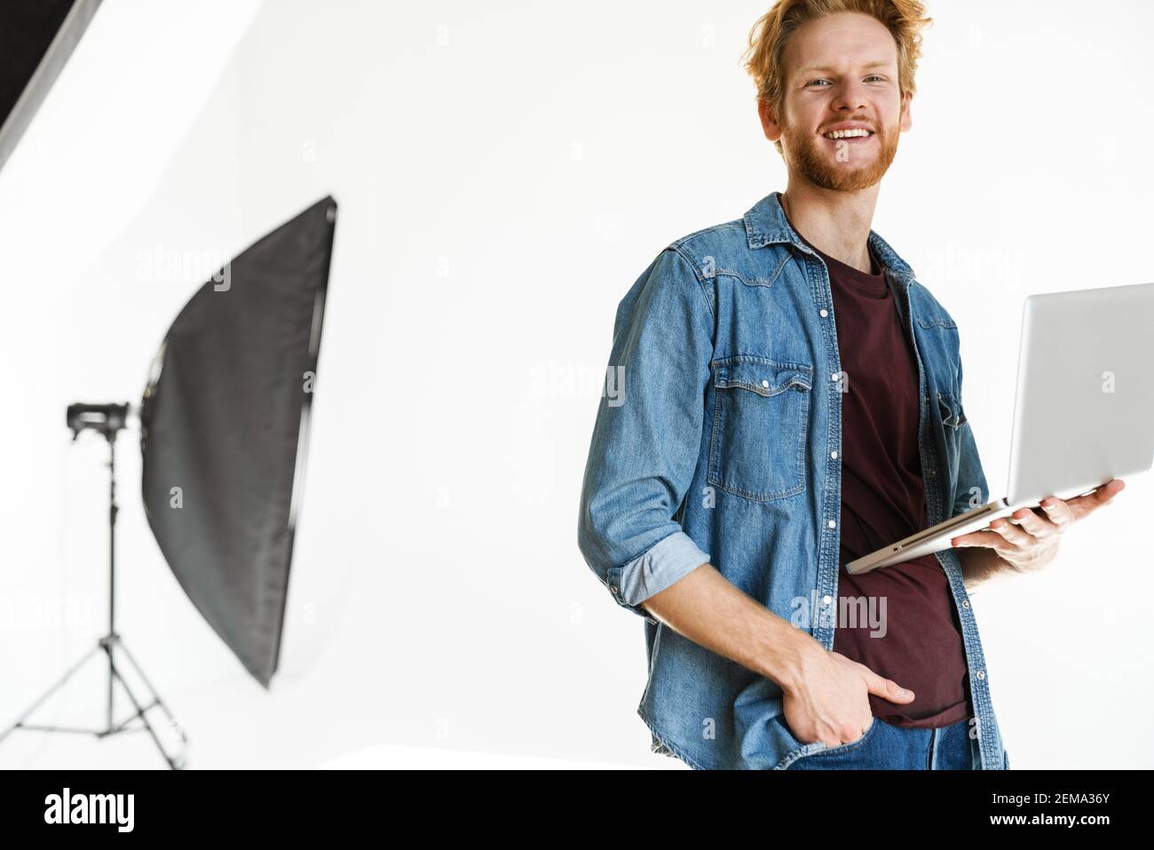 Happy handsome ginger guy smiling while working with laptop in studio ...