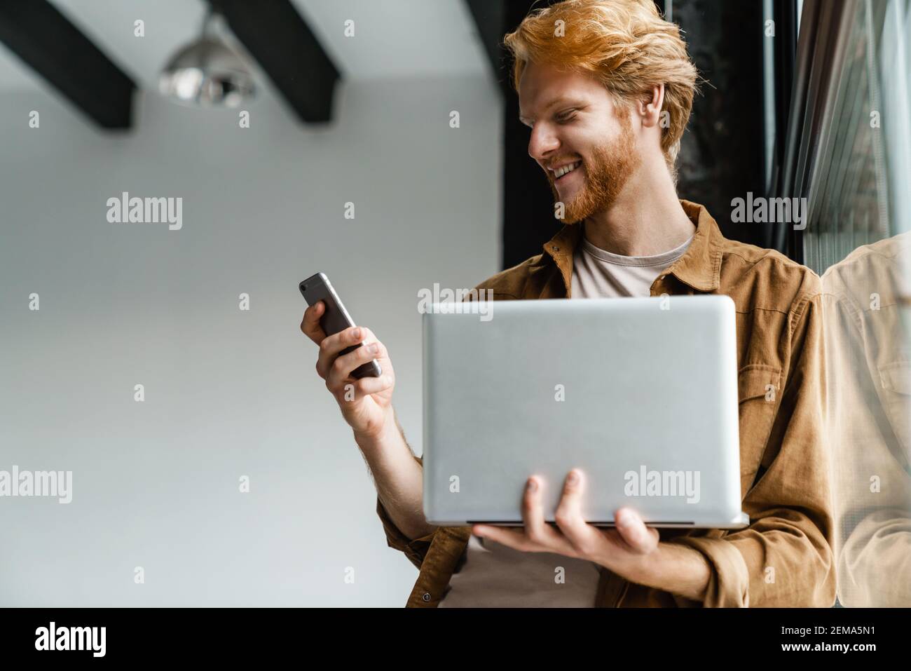 Joyful redhead guy smiling while working with laptop and cellphone near ...