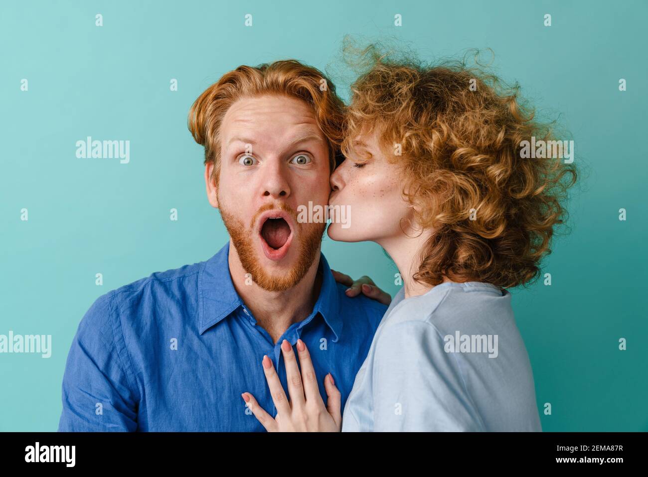 Beautiful young couple kissing on a cheek isolated over blue background ...