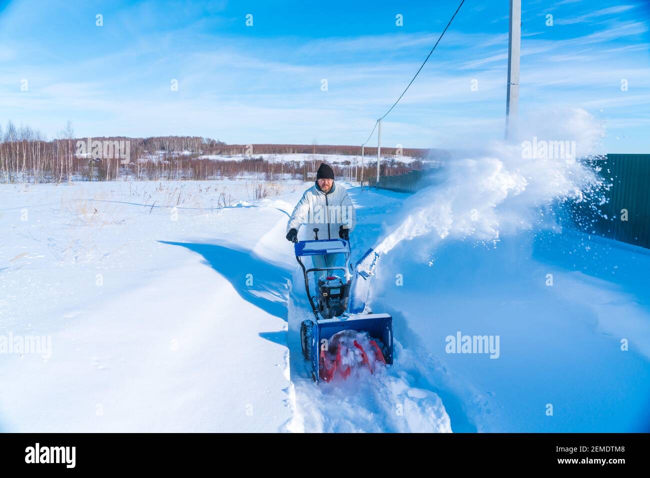 A man in a white jacket removes snow from a rural road with a ...