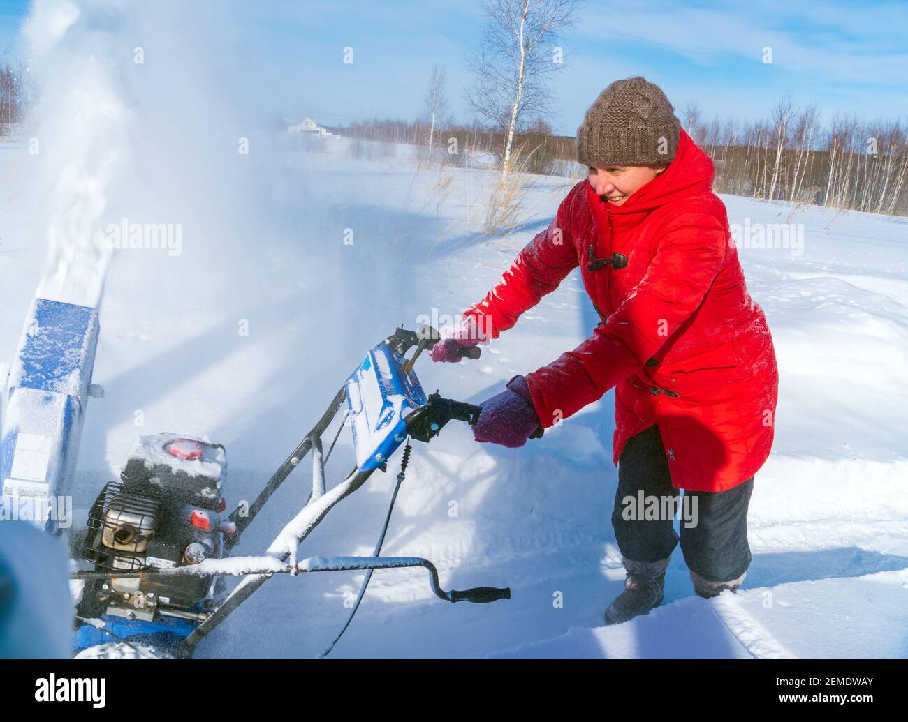 A woman in a red jacket removes snow from a rural road with a blue ...