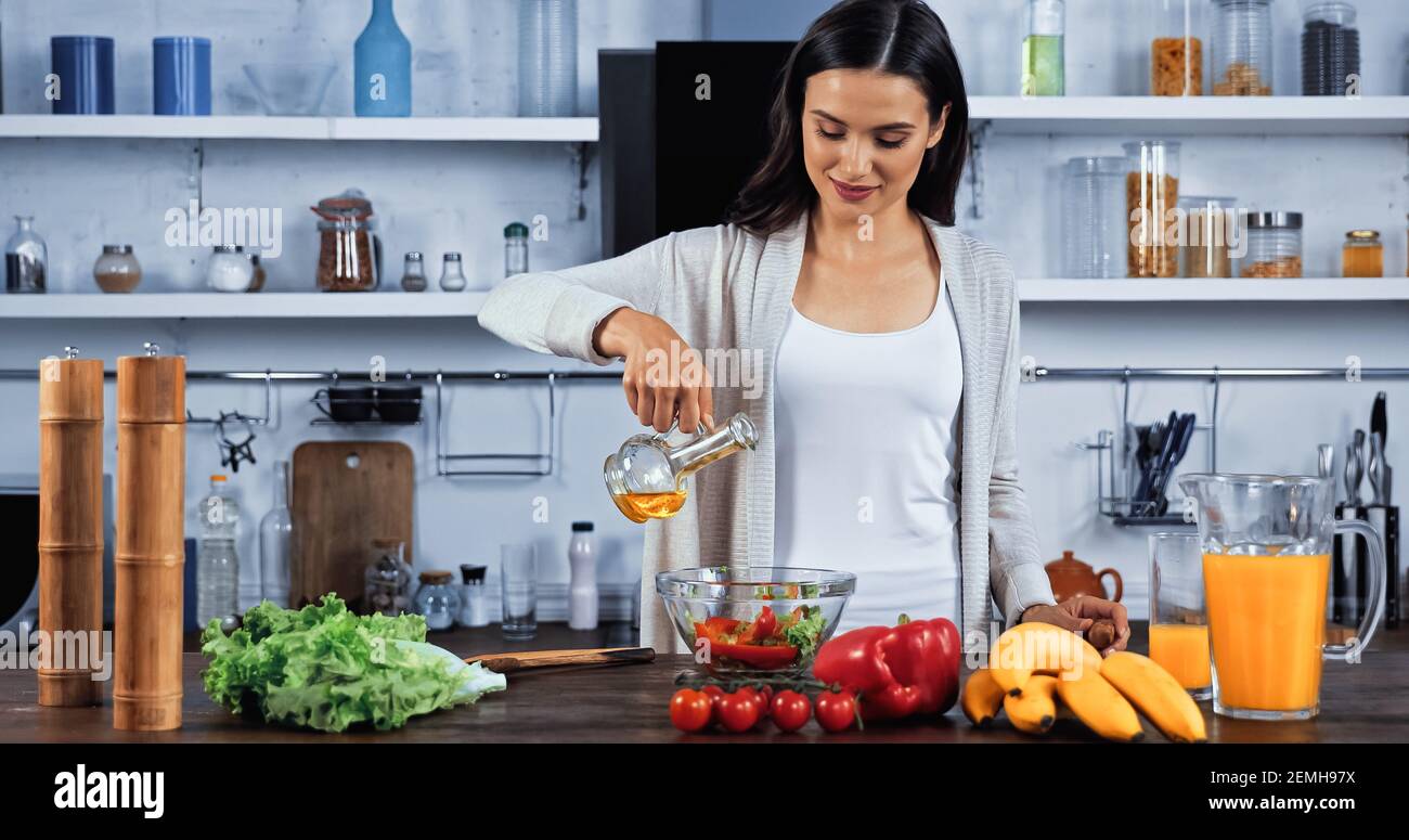 Brunette woman pouring oil in salad near fresh ingredients in kitchen ...