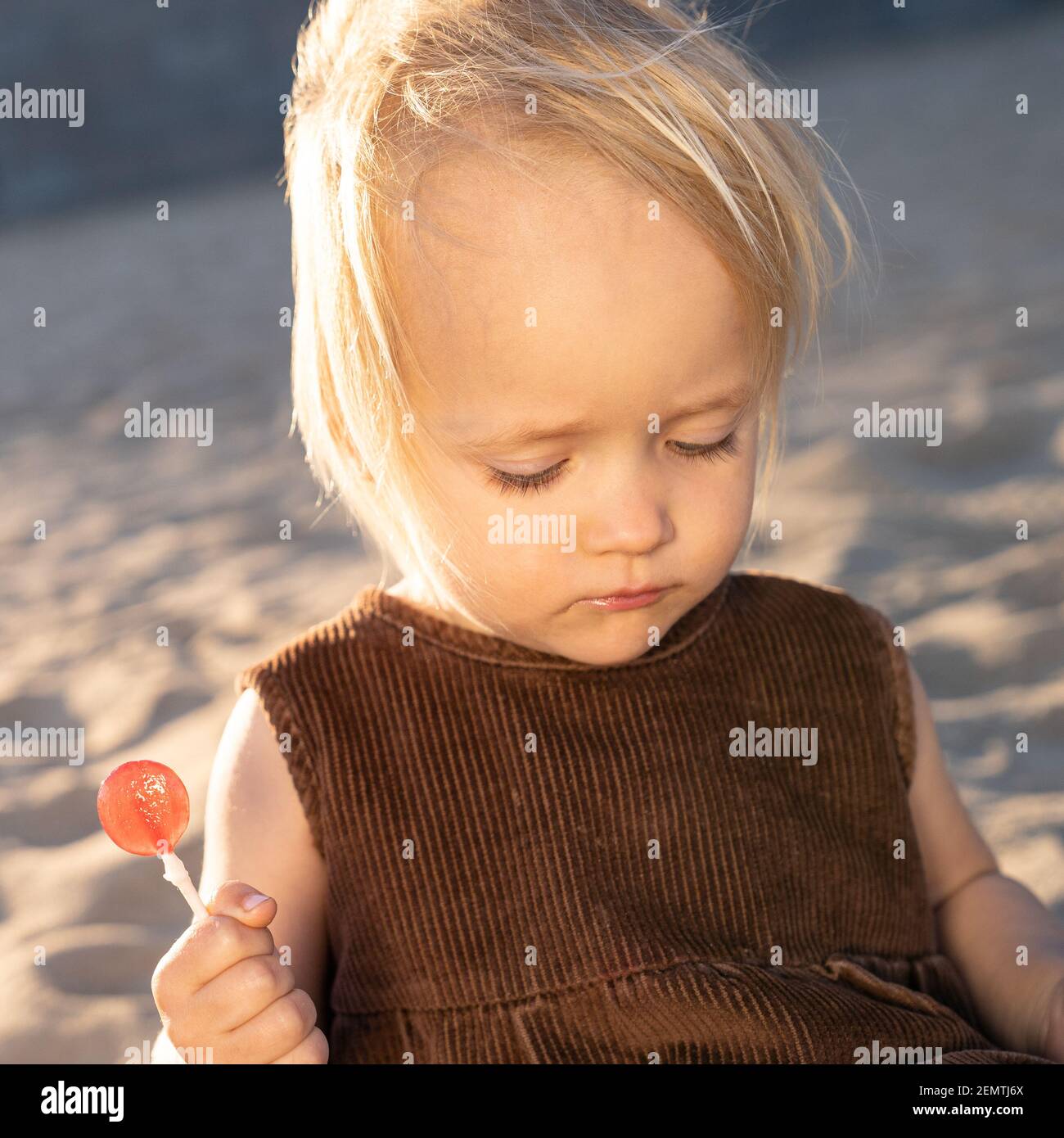 Pretty little child girl in brown dress looking down, touching lollipop ...