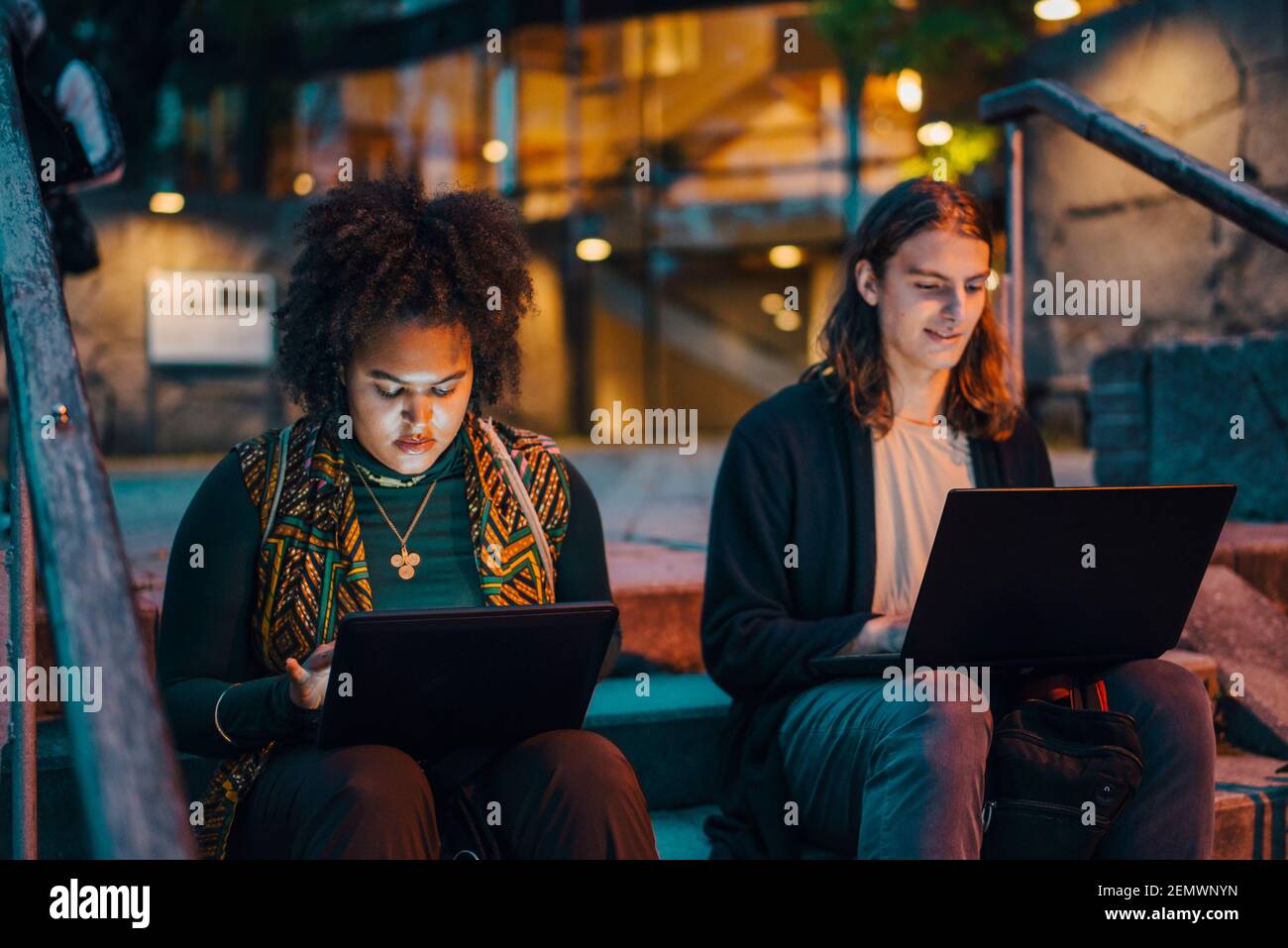 Young male and female students using laptop while sitting on steps in ...