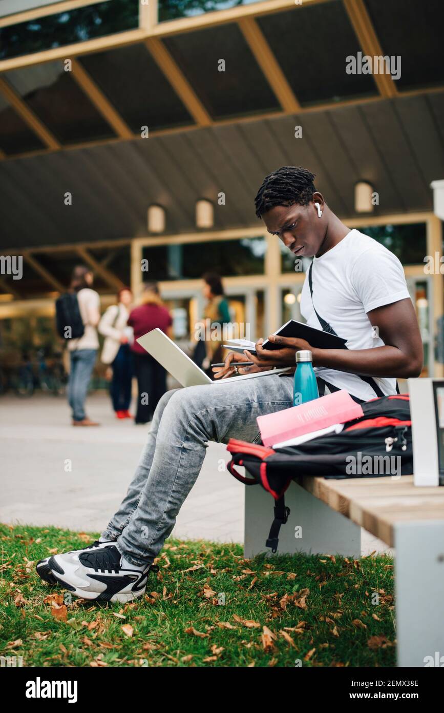 African male student reading book while sitting on bench in university ...