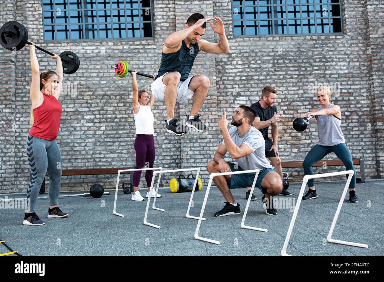 Young athlete jumping over hurdles while people around him lift weights ...