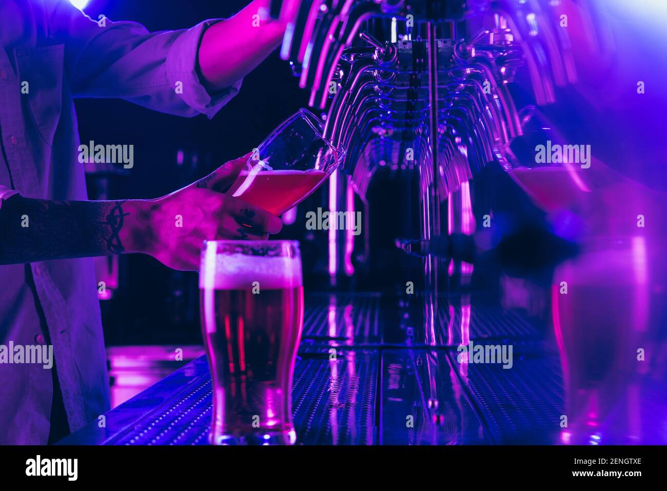 Young man bartender pouring dark beer from tap to glass in neon light ...