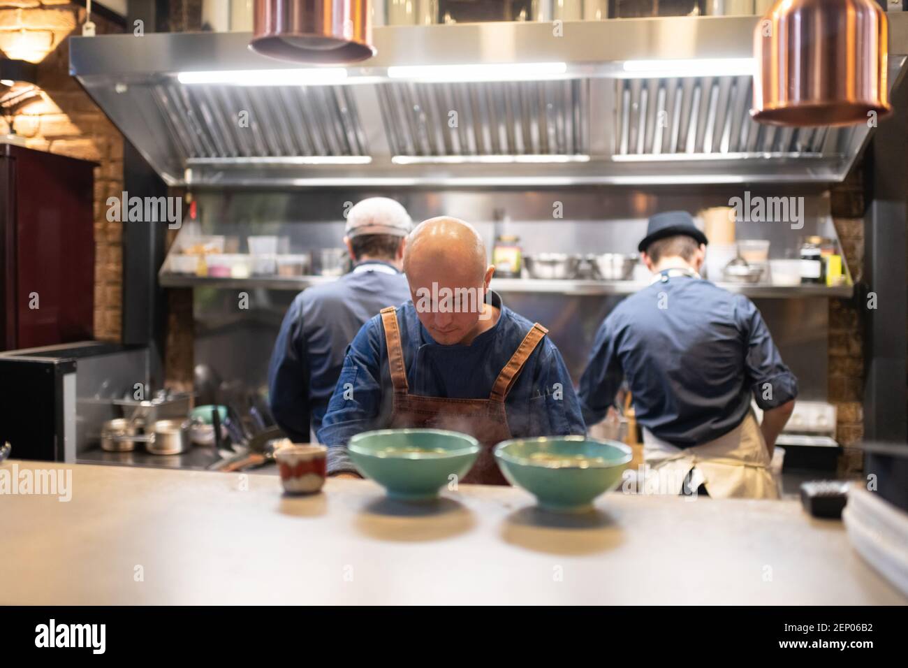 Bald man cooking food with coworkers behind counter with bowls of soup ...