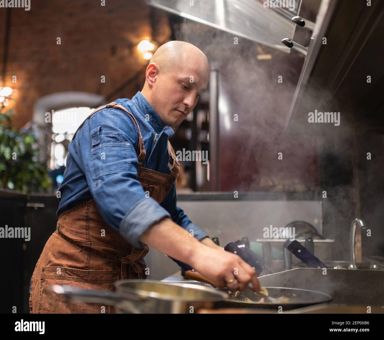 Bald man in apron frying tasty food on pan while working in kitchen of ...