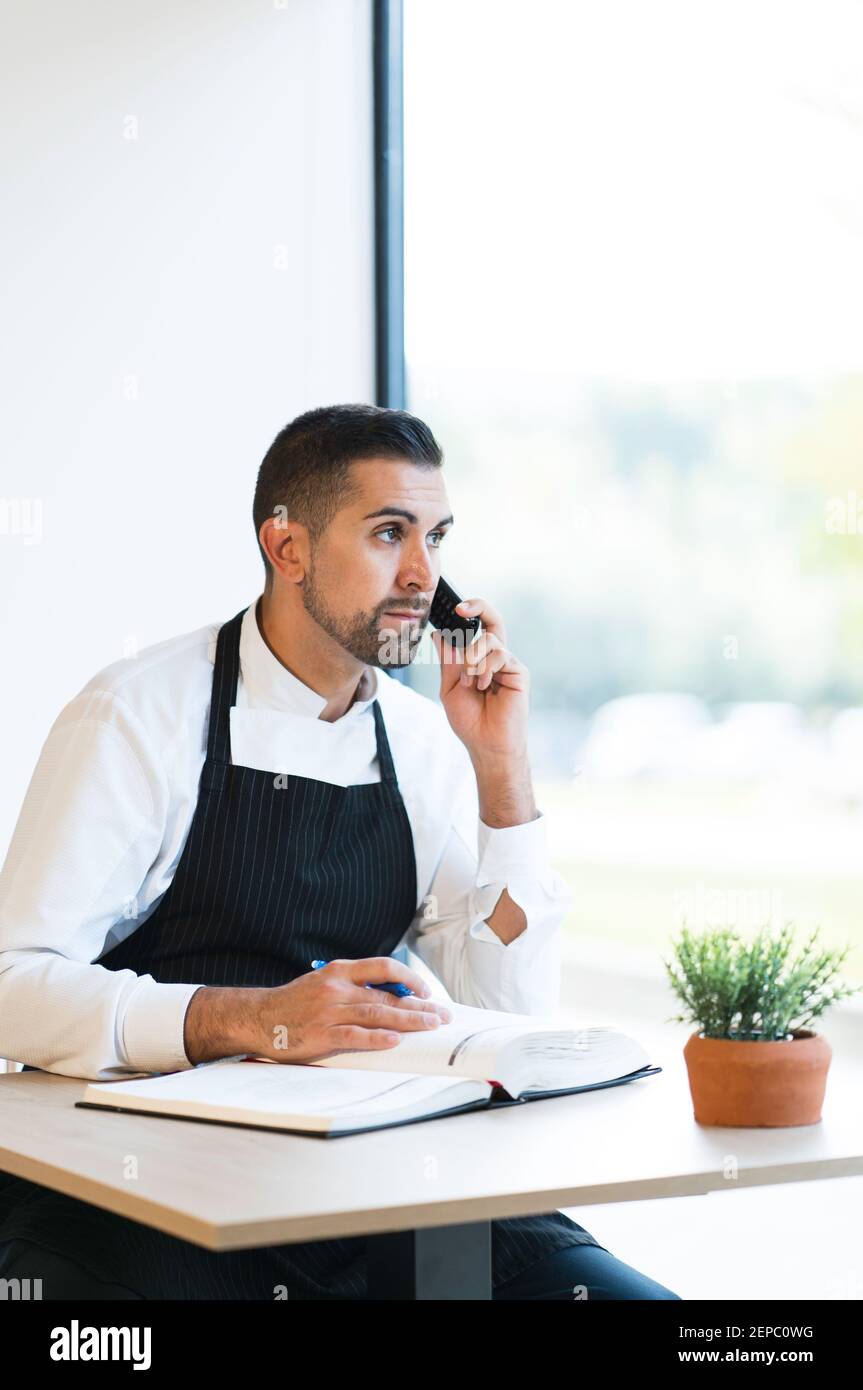 Young chef checking reservations talking by phone in restaurant wearing ...