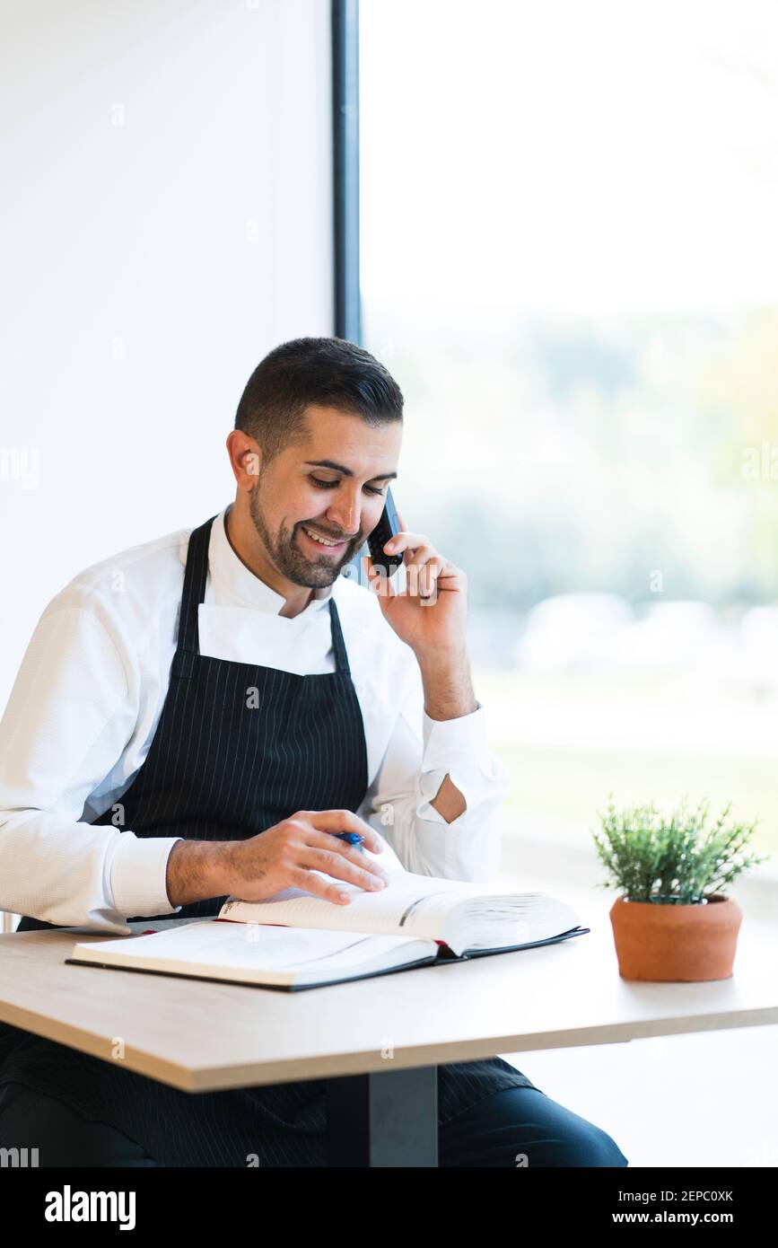 Young chef checking reservations talking by phone in restaurant wearing ...