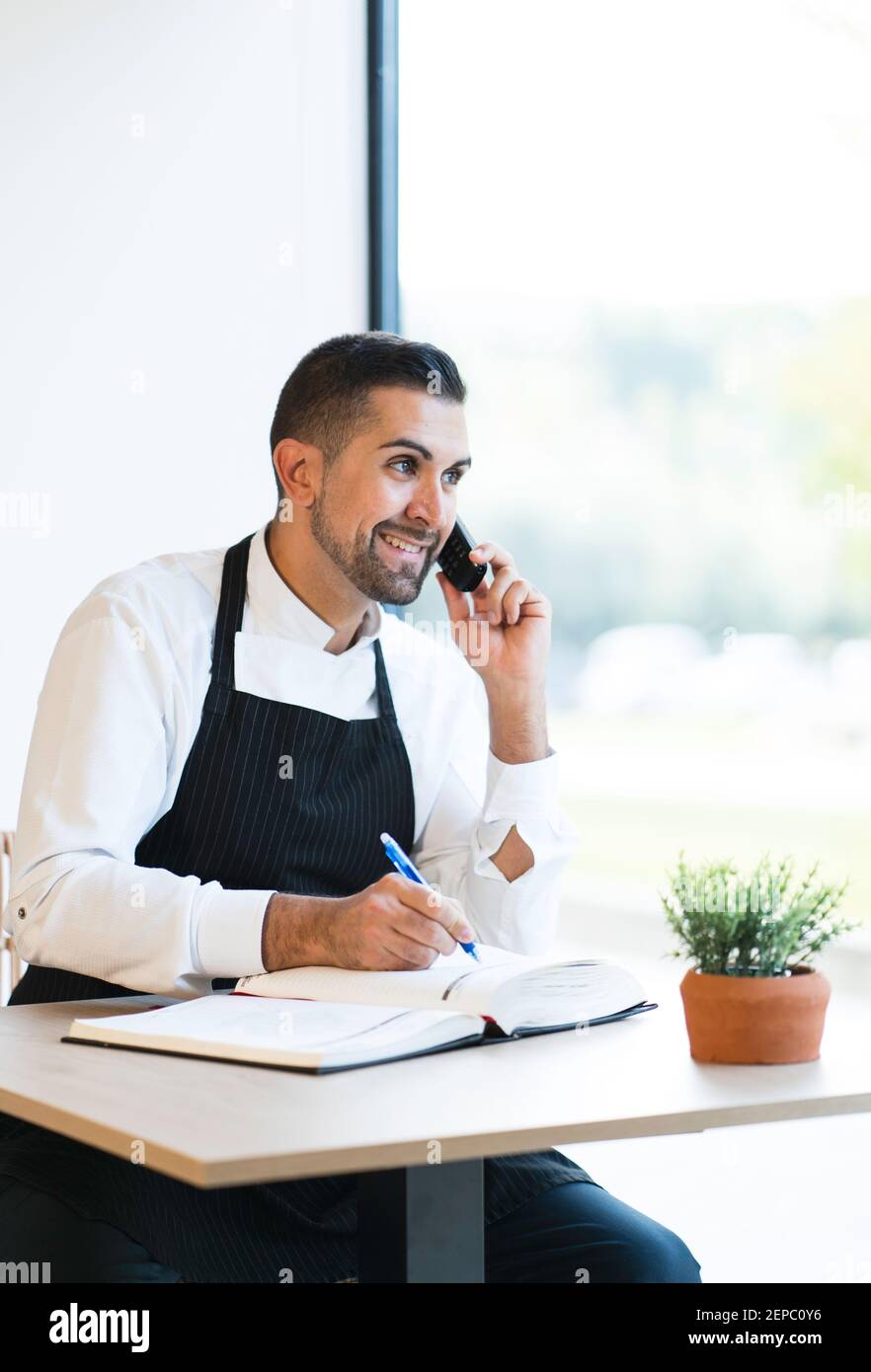 Young chef checking reservations talking by phone in restaurant wearing ...