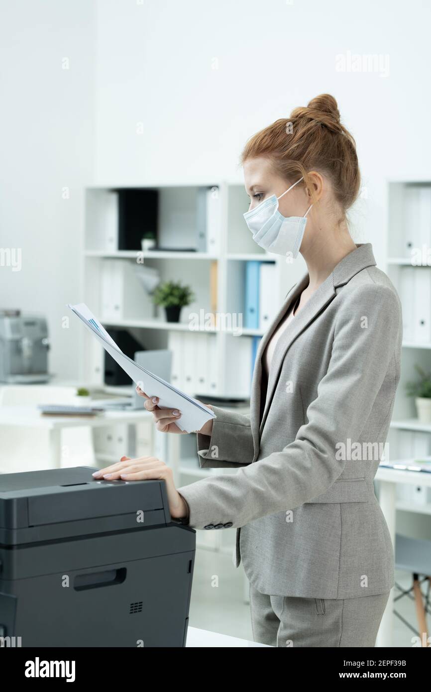 Young secretary in grey suit and protective mask looking through one of ...