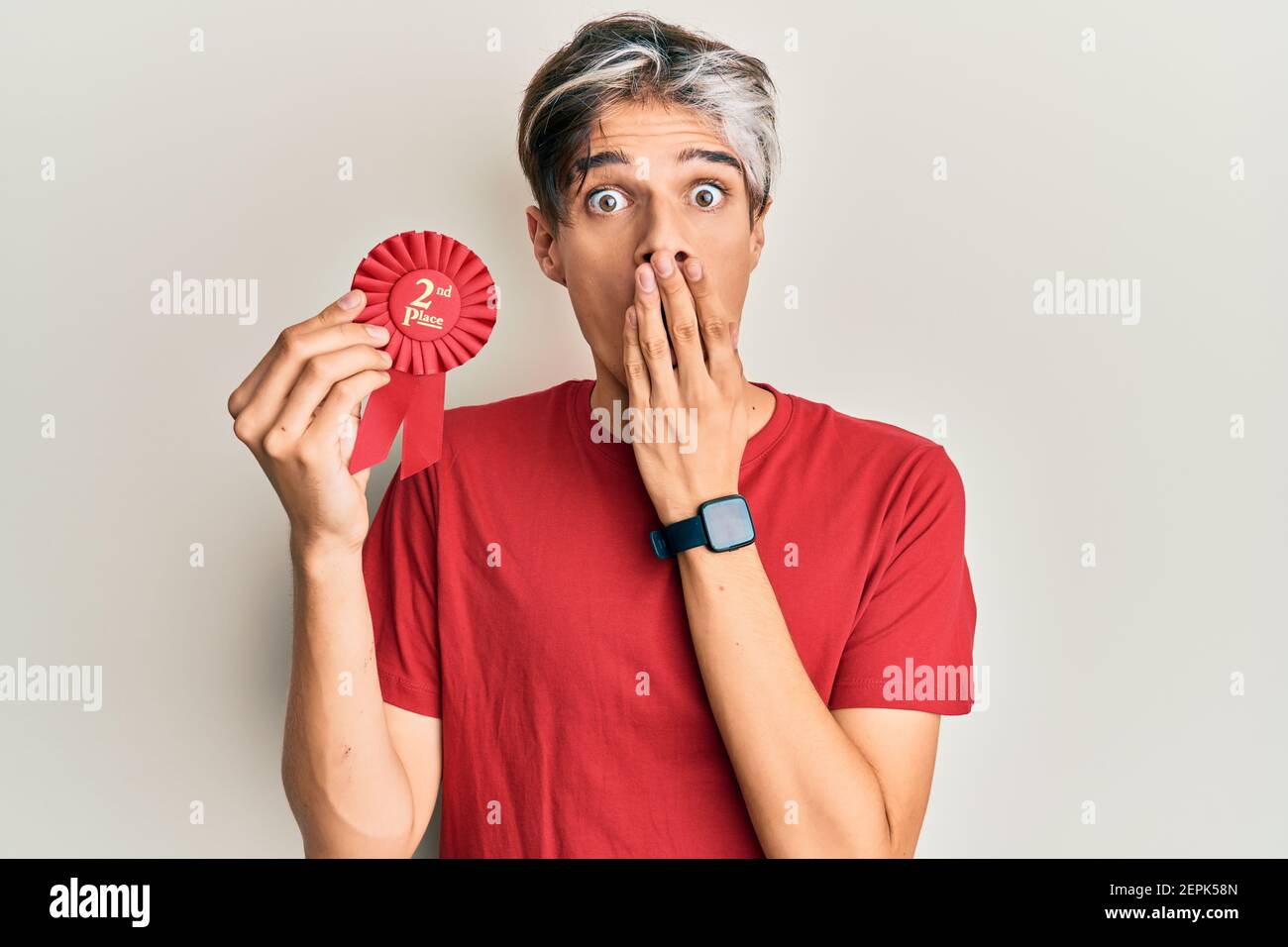 Young hispanic man holding second place badge covering mouth with hand ...