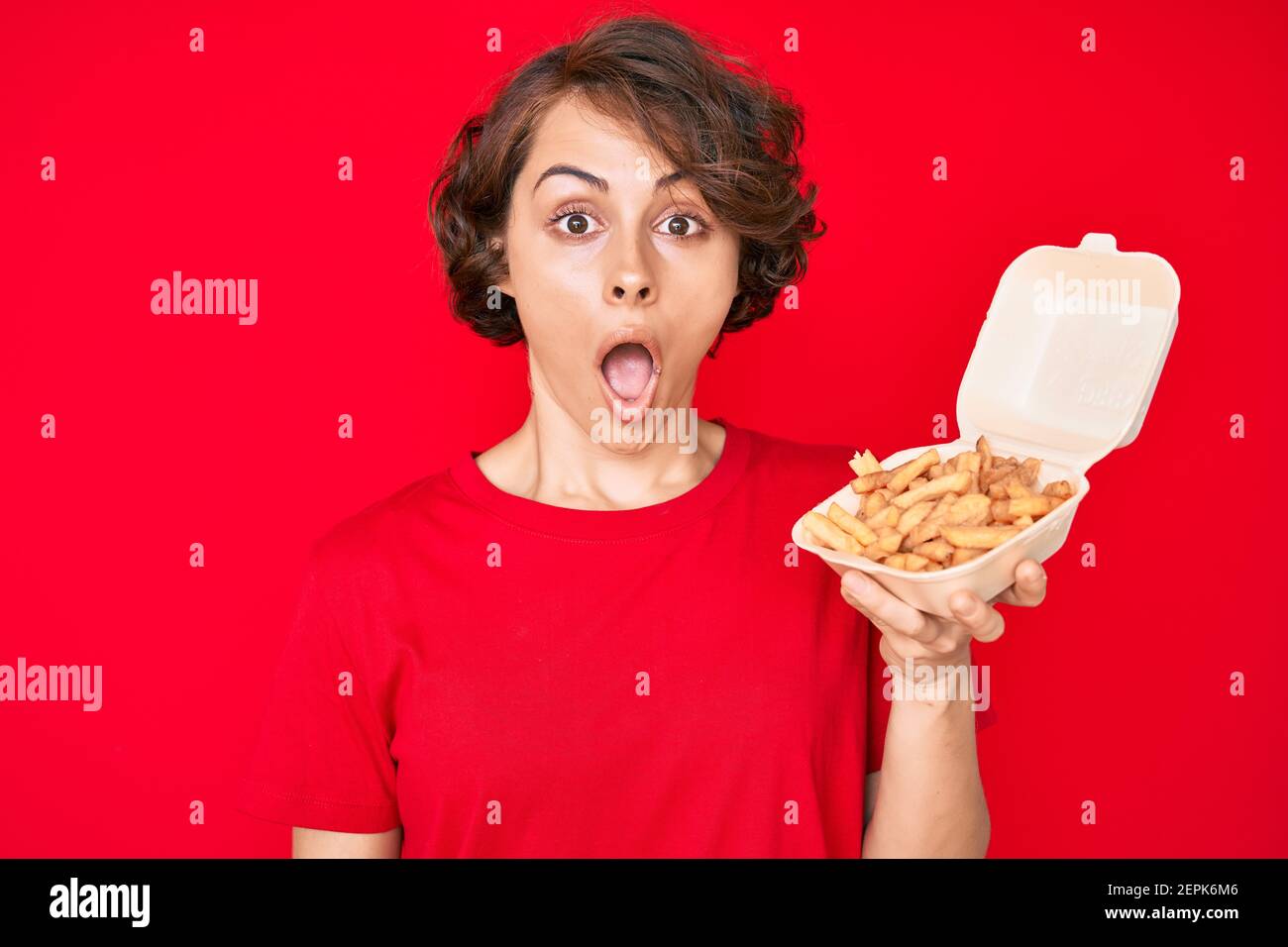 Young hispanic woman holding potato chip scared and amazed with open ...