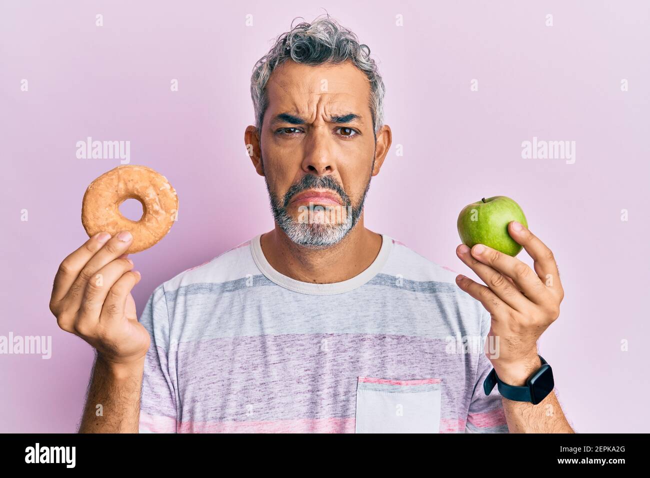 Middle age grey-haired man holding green apple and donut depressed and ...